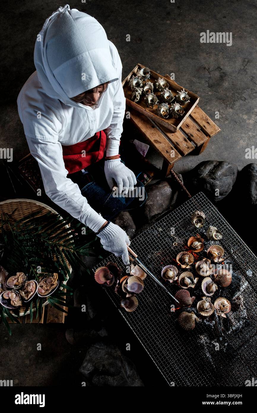 Japanese Ama diver cooks shellfish on grill in traditional Ama hut ...