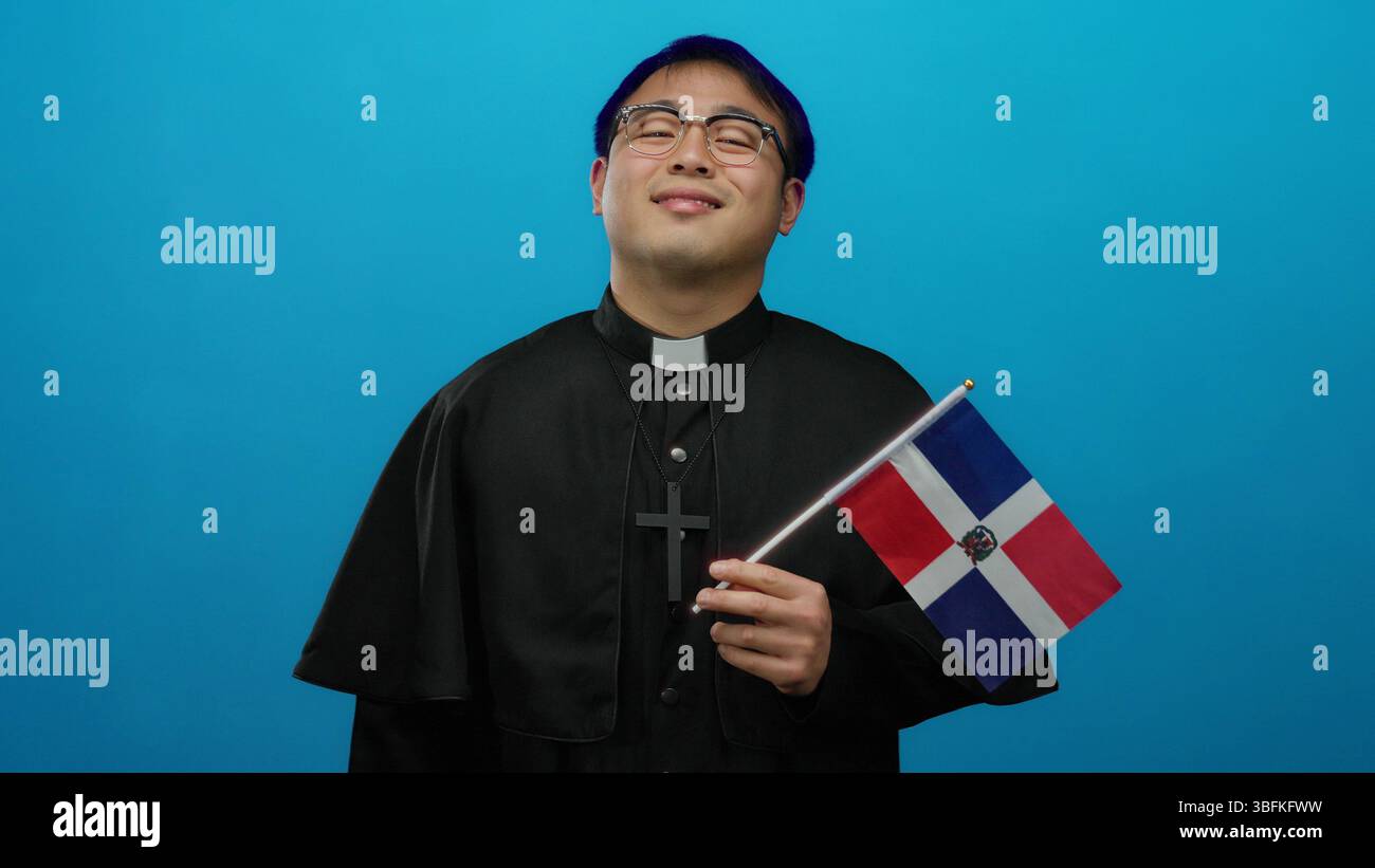 Priest holding dominican republic flag smiling against blue isolated ...