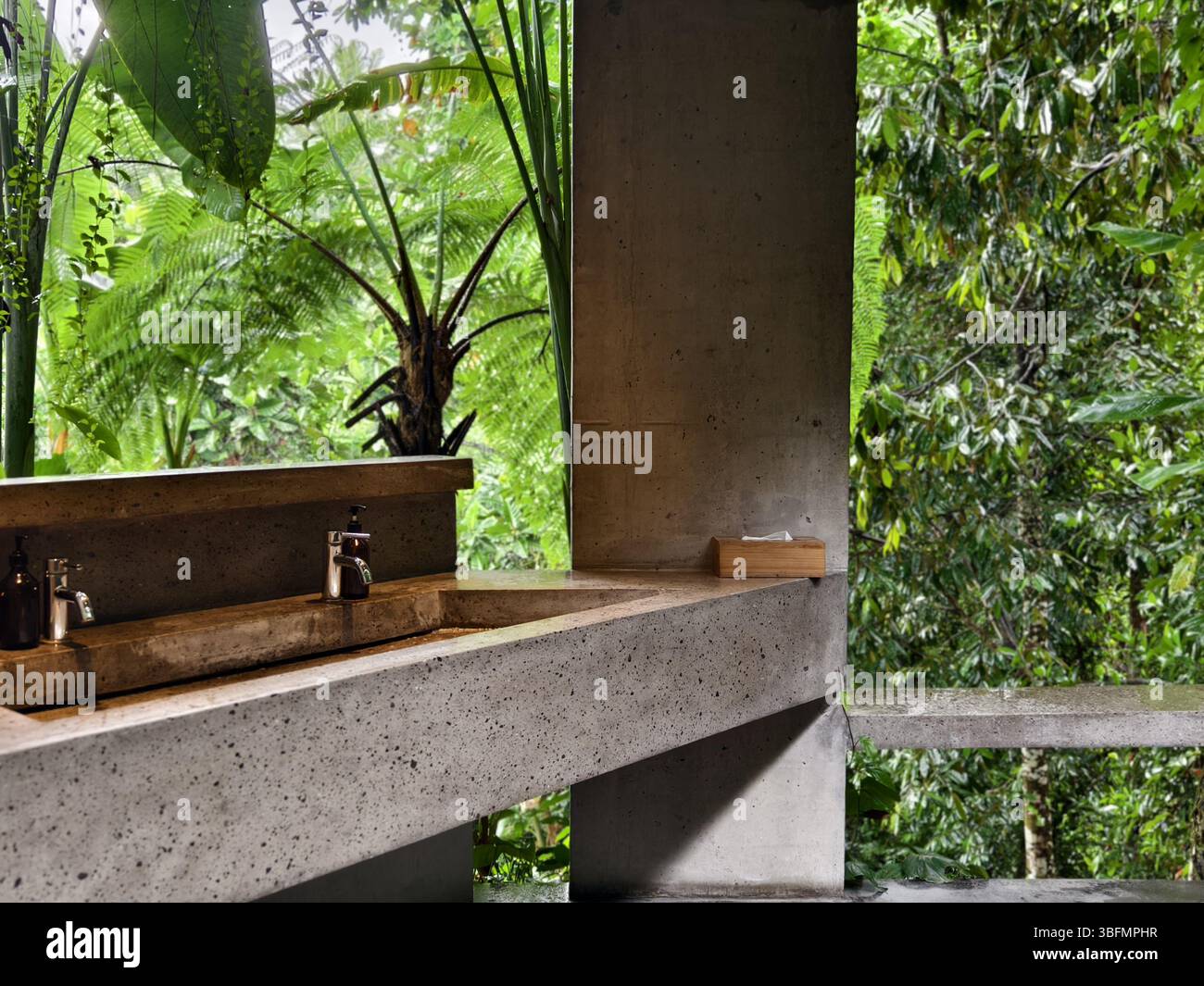An open-air concrete sink station with two faucets next to a lush green ...