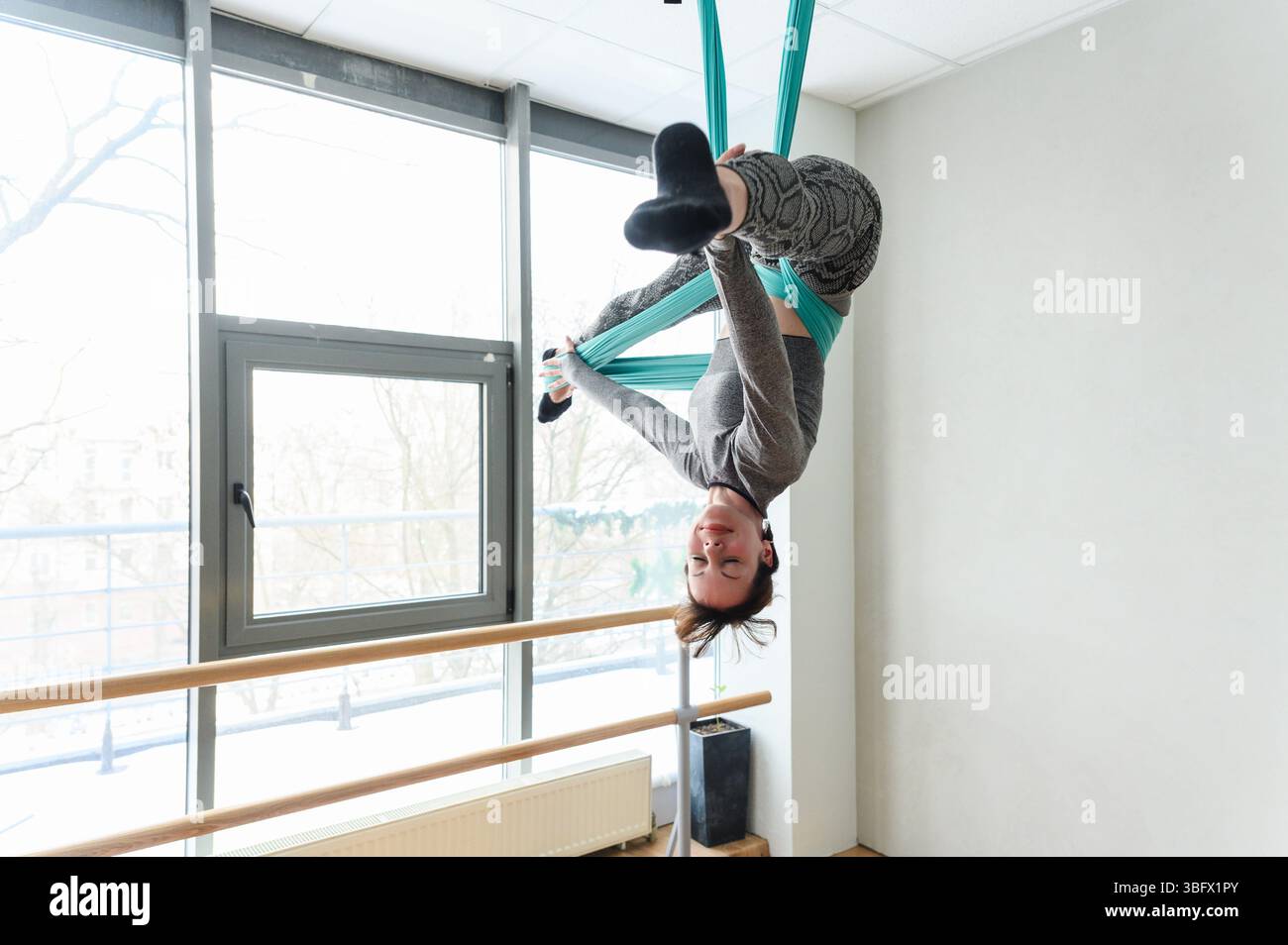 Athletic woman performing an inverted pose on teal aerial silks, legs stretched and body ...
