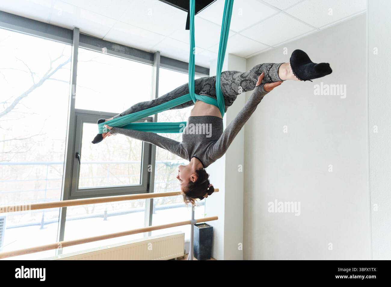 Athletic woman performing an inverted pose on teal aerial silks, legs ...