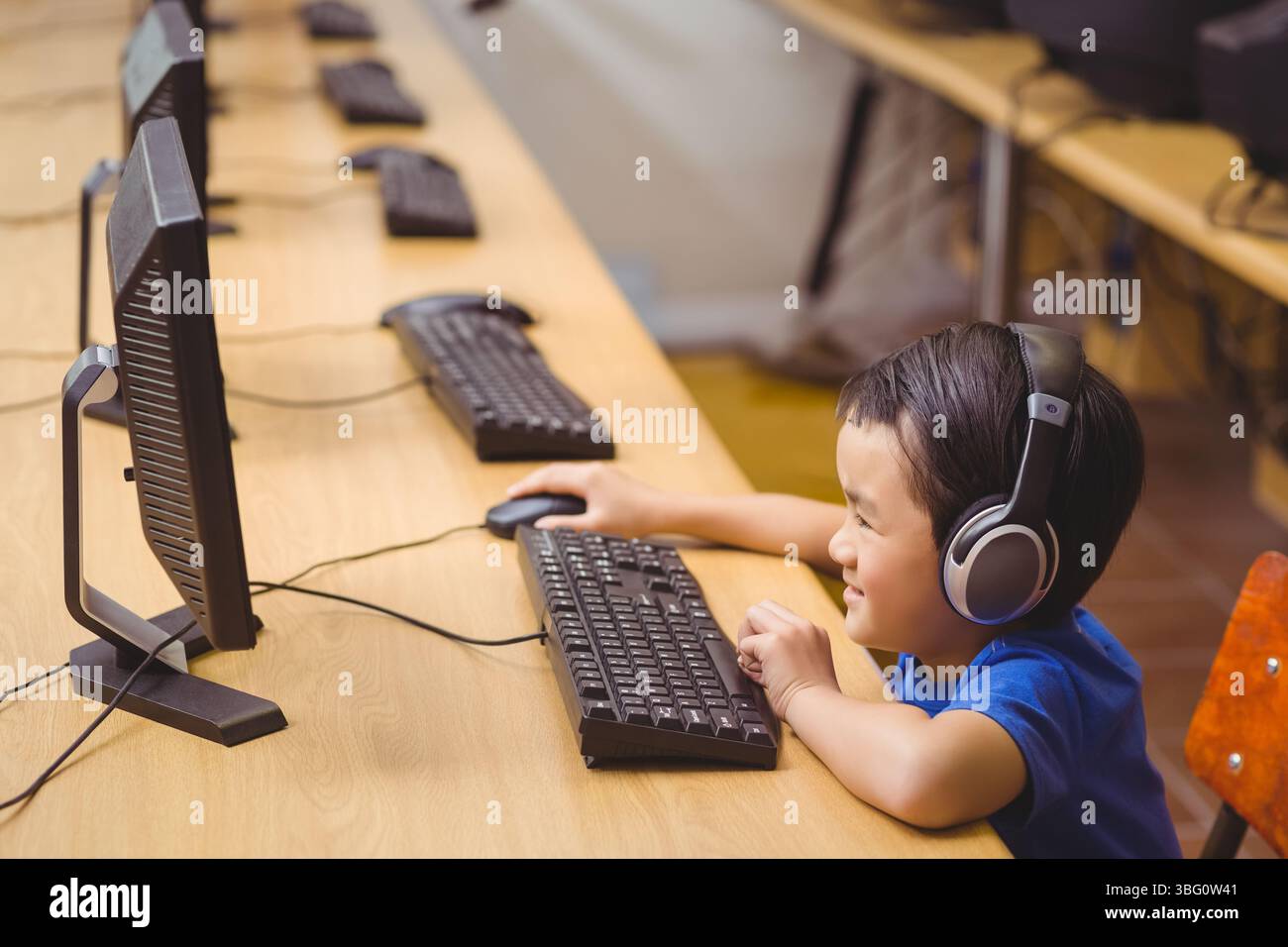 Asian boy wearing headphones reaching for mouse at desk with monitor in ...