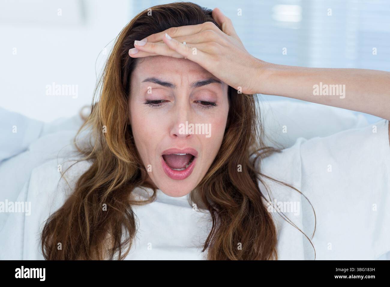 Woman pressing hand against forehead and gasping on white sofa with ...