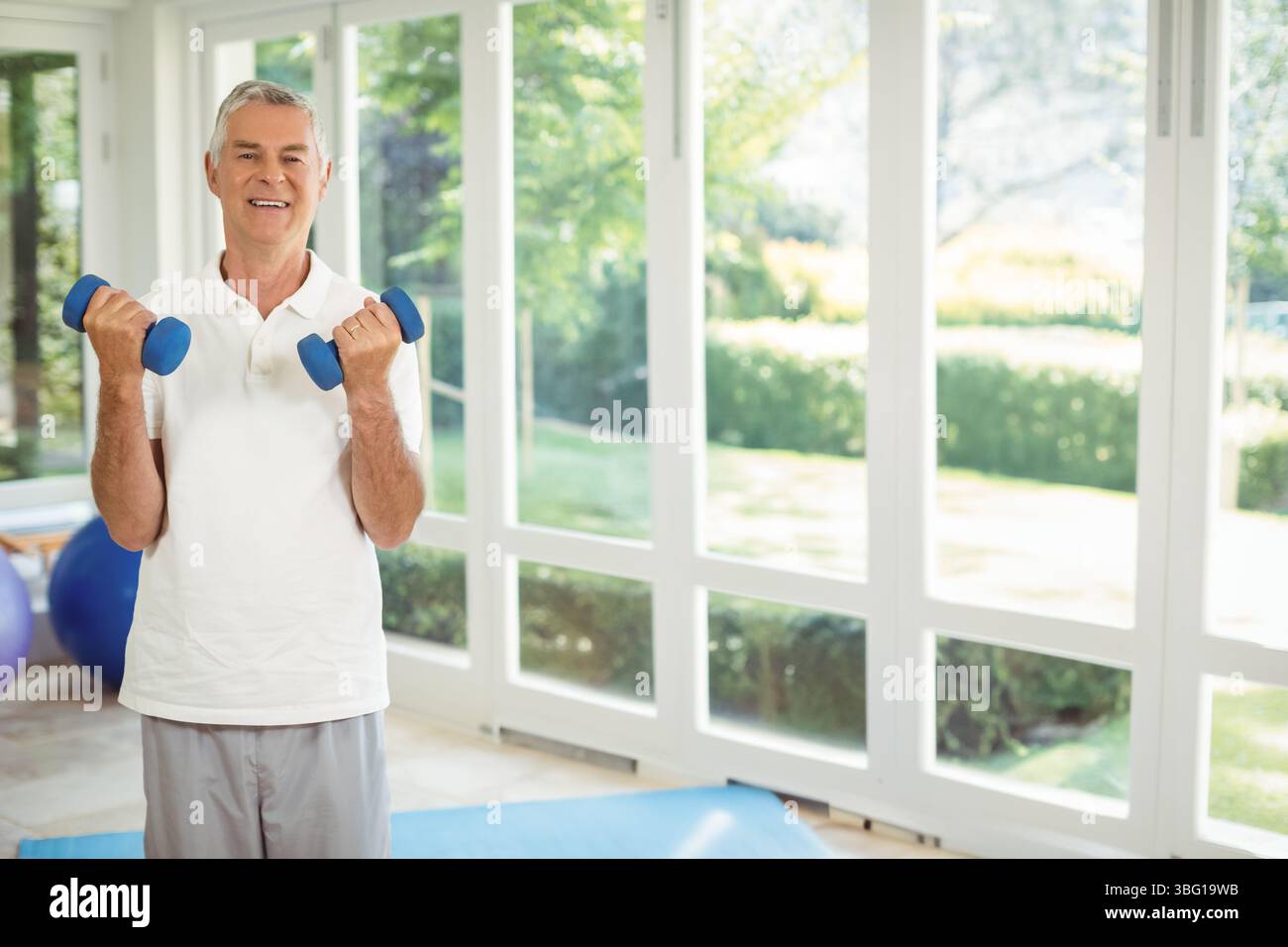Senior man standing in sunroom lifting blue dumbbells by windows with ...