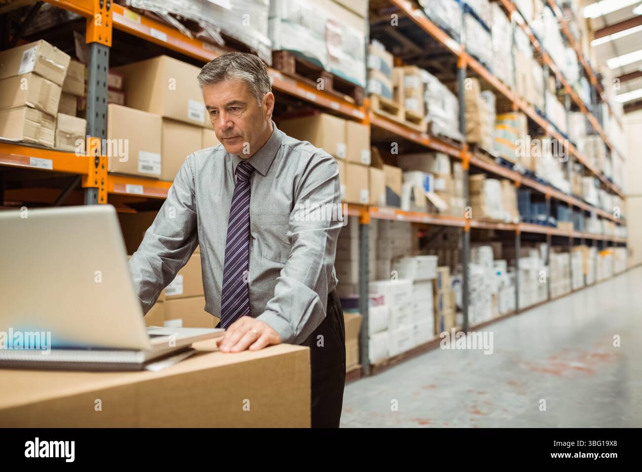 Man in his 50s wearing tie typing on laptop at box in warehouse aisle ...