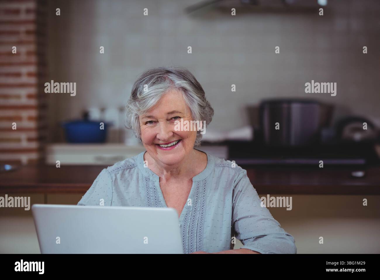 Senior woman typing on laptop at kitchen counter with blue pot and ...