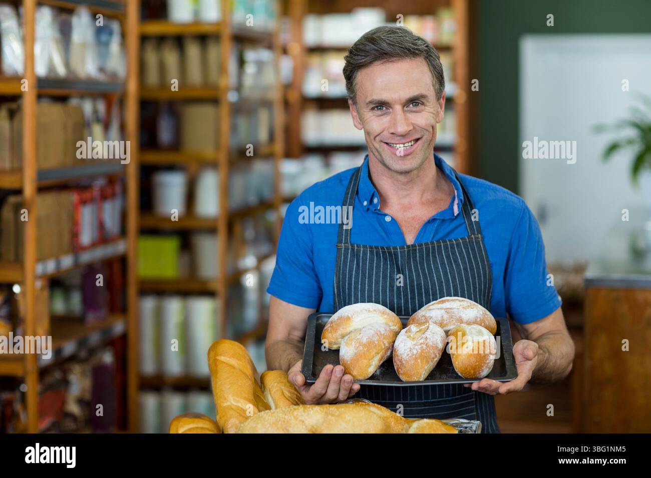 Mature male baker in dark apron presenting tray of four flour-dusted ...
