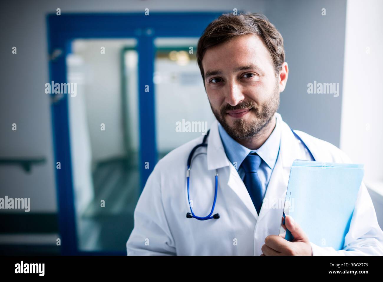 Male doctor standing in hospital hall wearing white coat holding folder ...