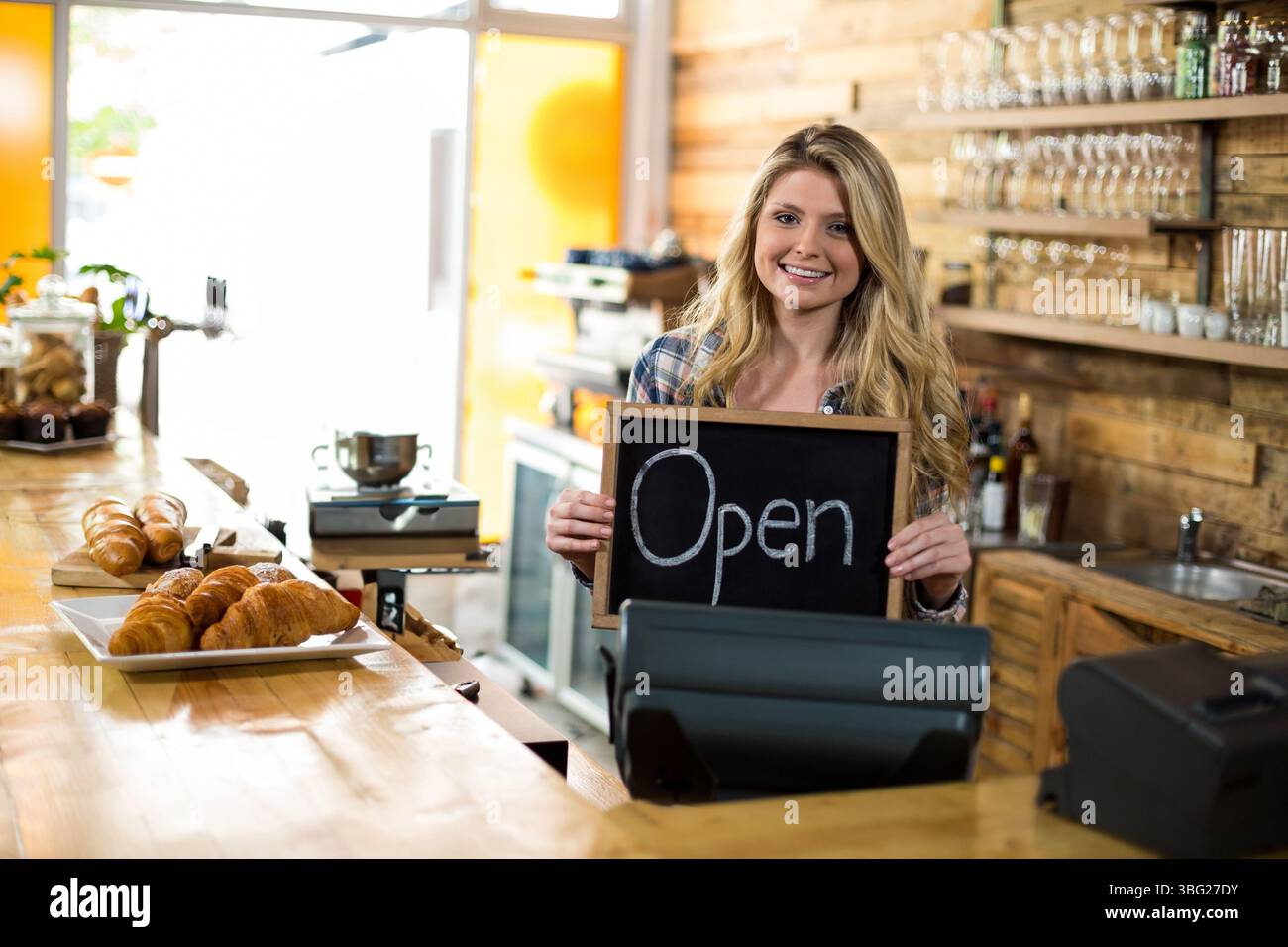 Blonde woman standing behind counter in bakery cafe holding Open ...