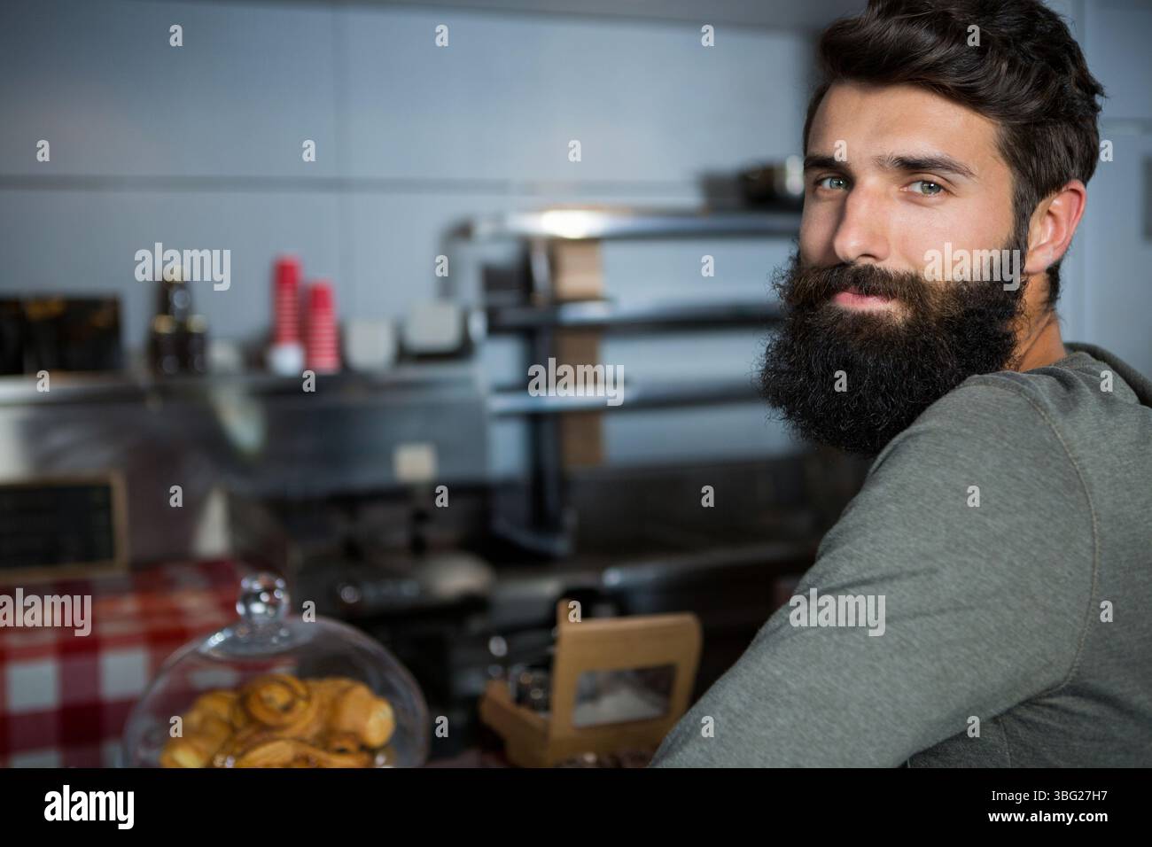 Bearded man sitting at café counter smiling with pastries under glass ...