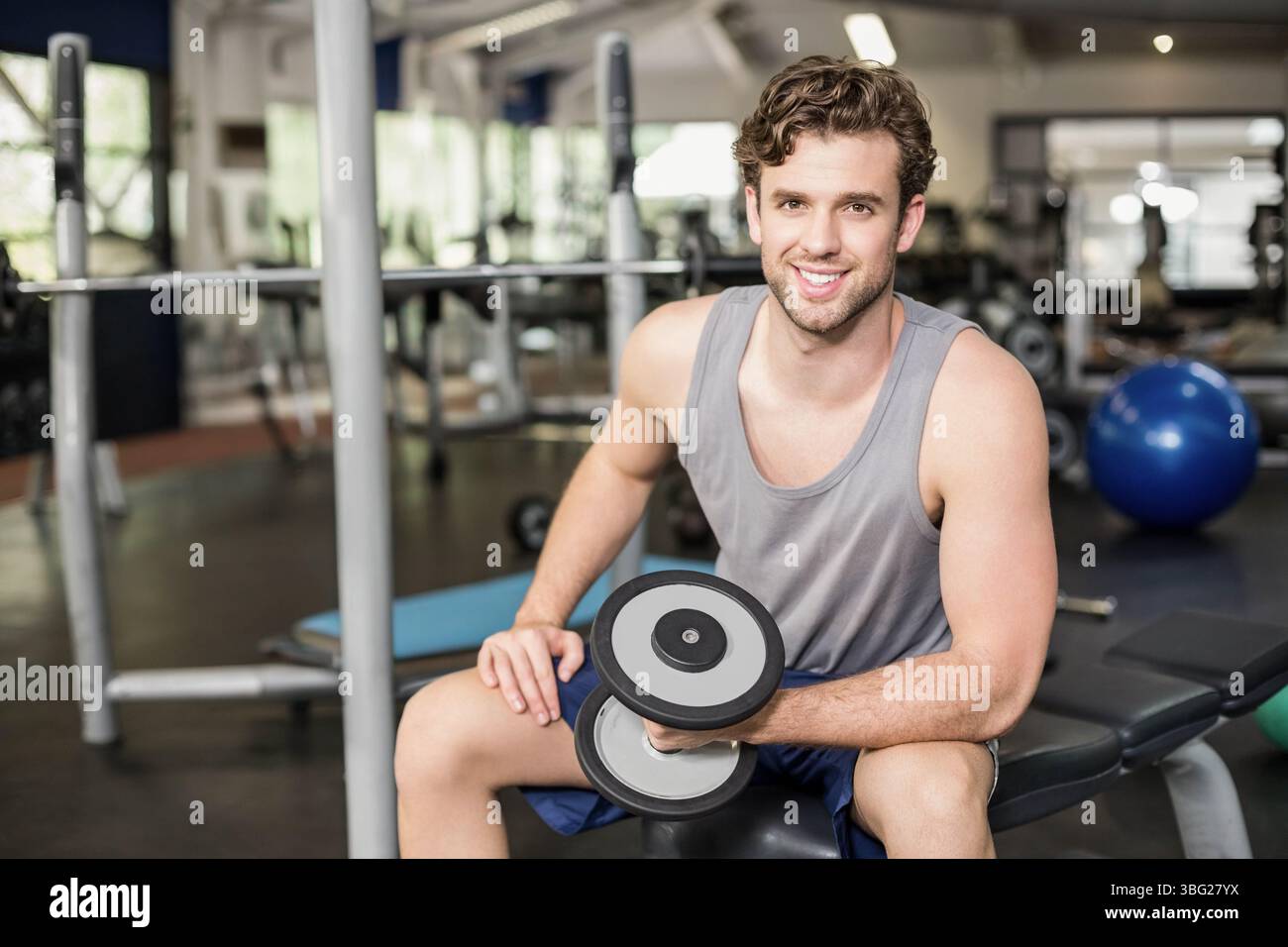 Fit man lifting dumbbell while sitting on weight bench at gym with ...