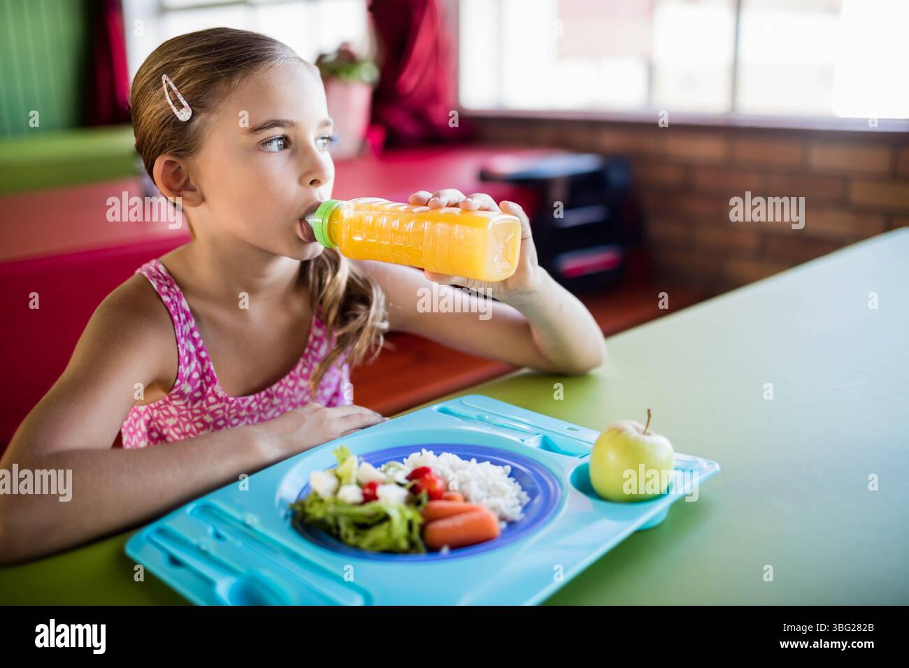 Girl sipping orange juice from bottle in cafeteria, holding blue tray ...