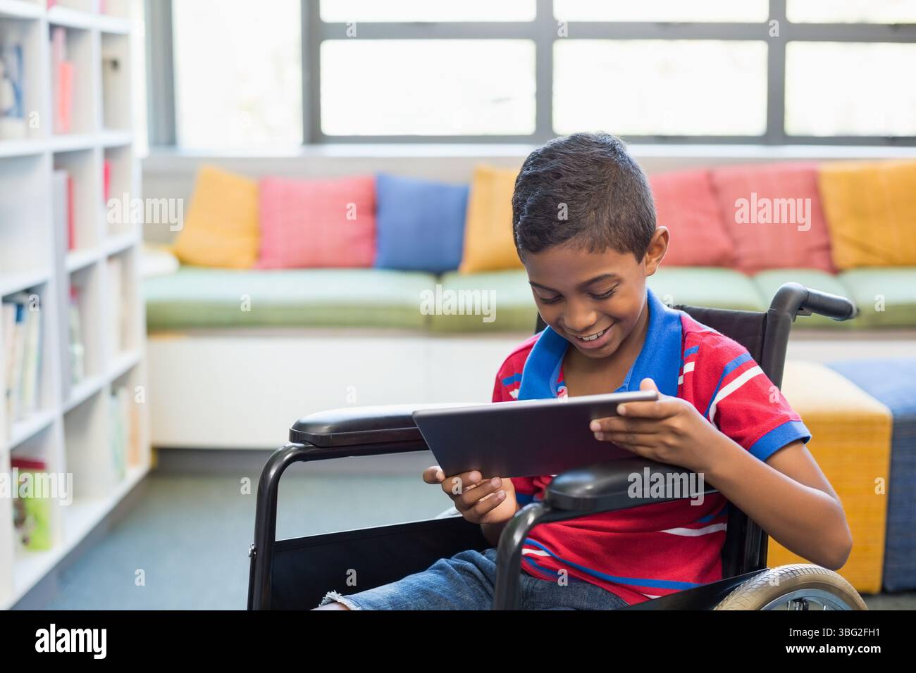 Boy is sitting in wheelchair, reading on tablet in library area with ...