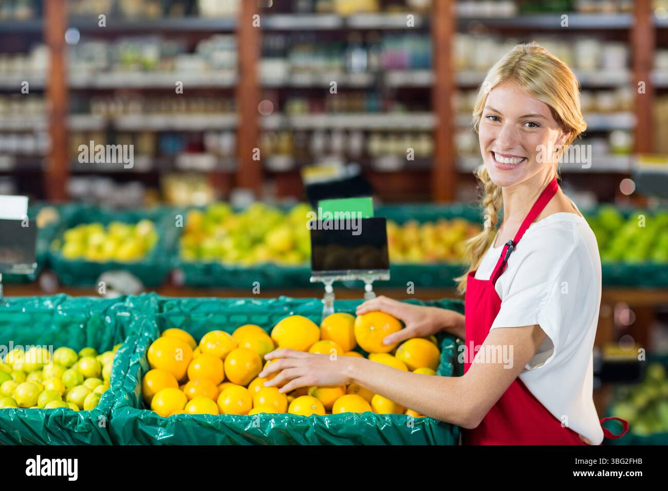 Female store employee in red apron arranging oranges in green bins at ...