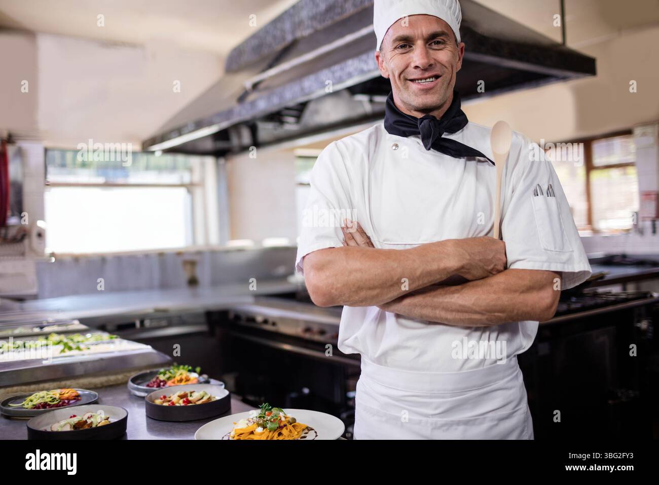 Male chef holding spoon under arm in commercial kitchen displaying ...