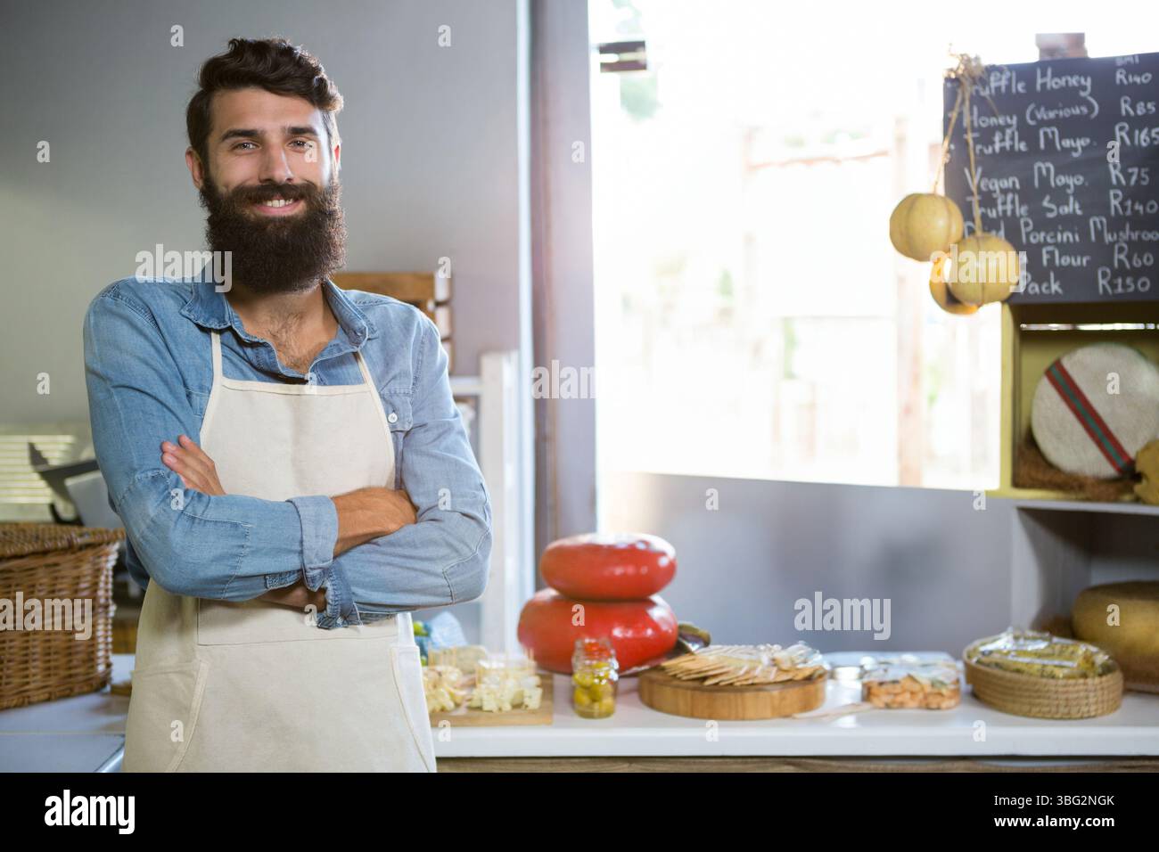 Male shopkeeper in apron smiling and showing cheese wheels and olives ...