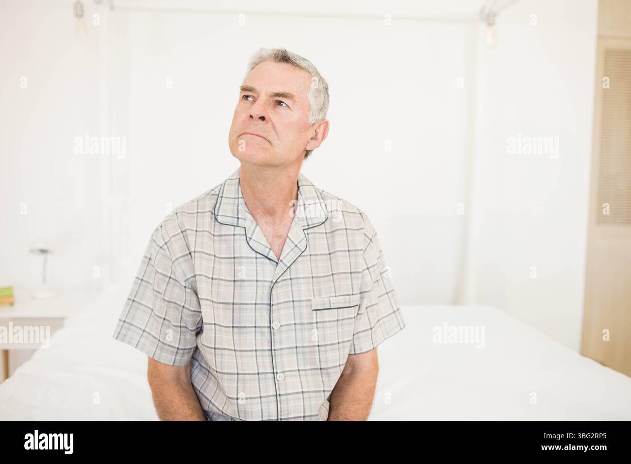 Senior man wearing pajamas sitting on bed edge in bedroom gazing upward ...