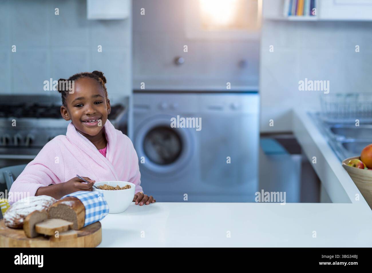African American girl eating cereal at kitchen counter in bathrobe beside cutting board, copy ...