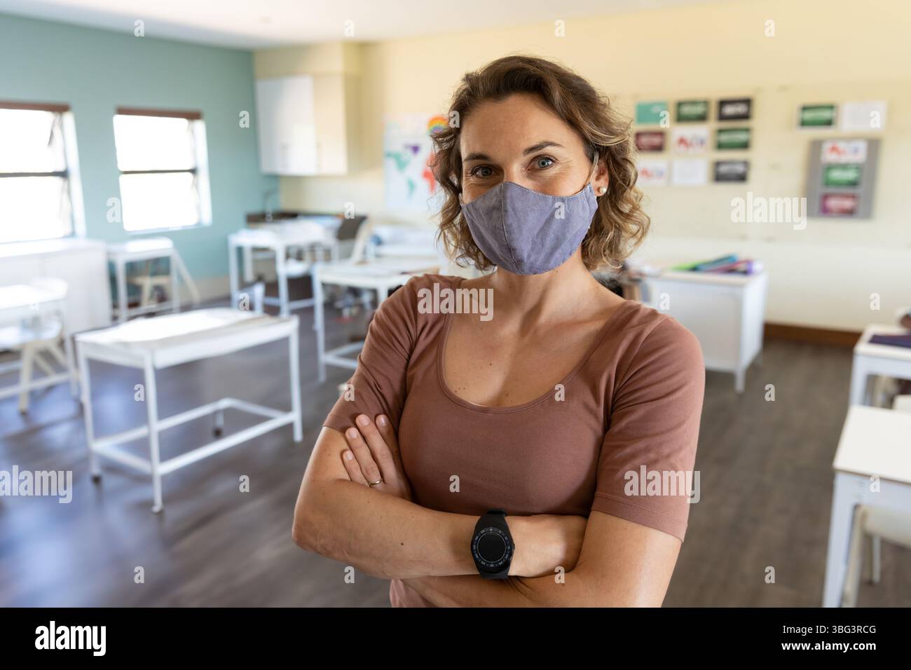 Female teacher standing with arms crossed in classroom wearing face ...