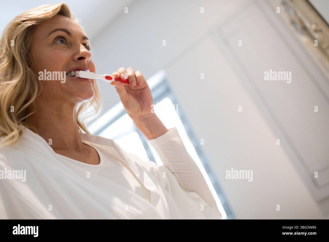 Mid adult woman brushing teeth with red white toothbrush in bathroom ...