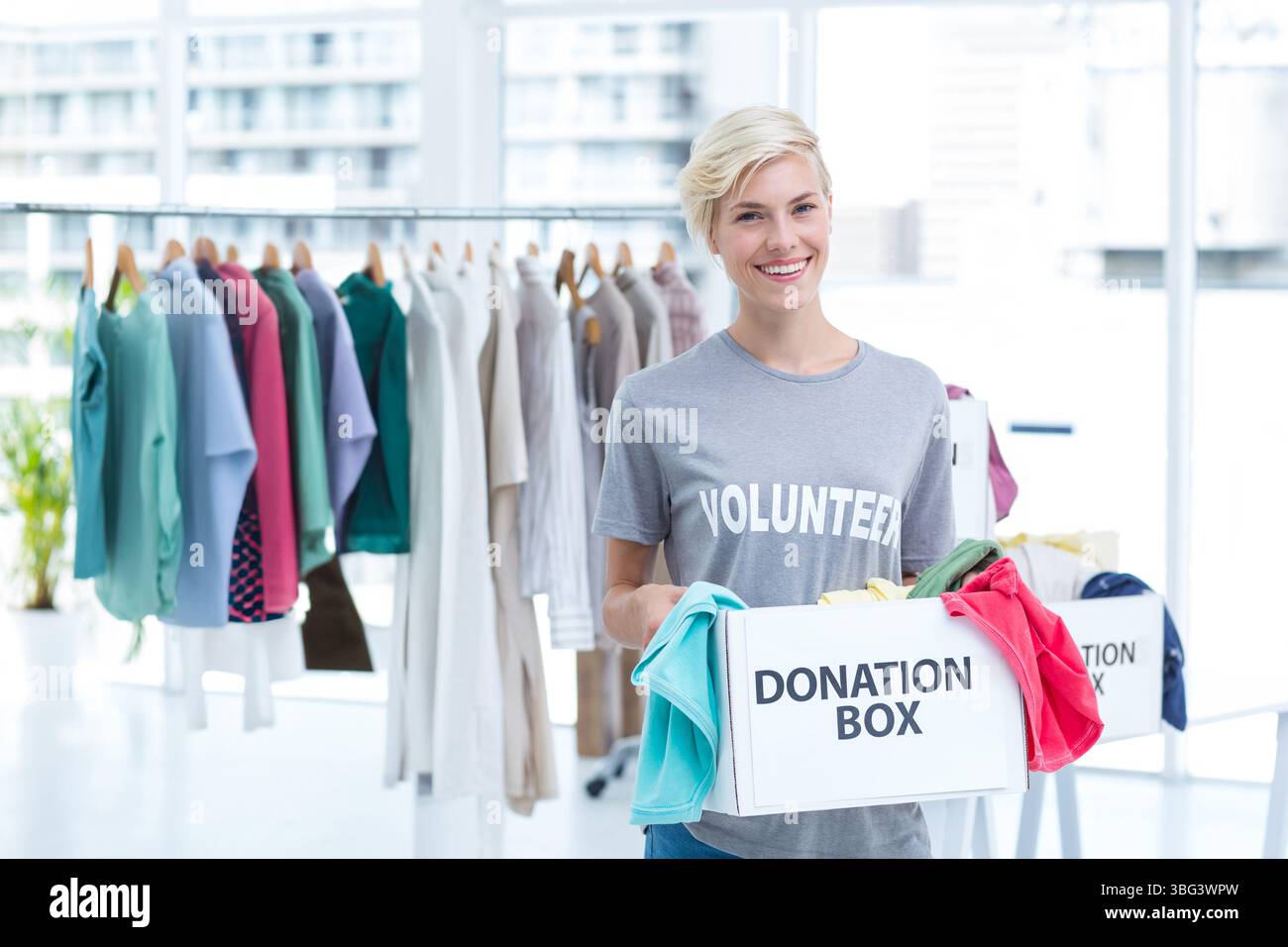 Smiling female volunteer holding donation box and standing by clothing rack in donation center ...
