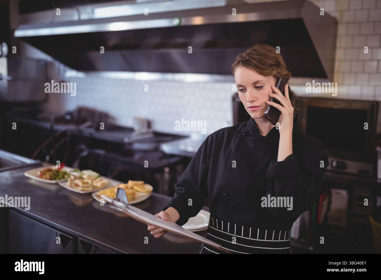 Female chef in chef coat holding smartphone reviewing order sheet at ...