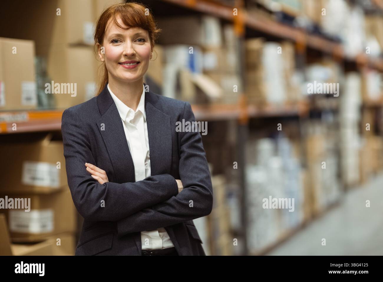 Female warehouse supervisor standing in warehouse aisle surveying pallet racks and labeled boxes ...