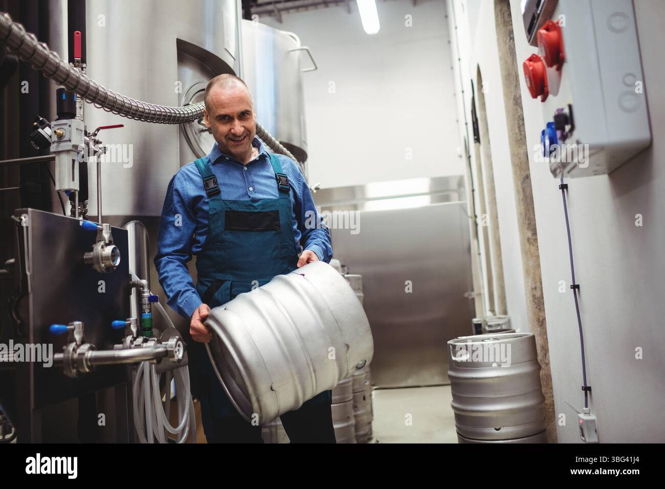 Male brewery worker wearing overalls lifting keg near stainless steel ...