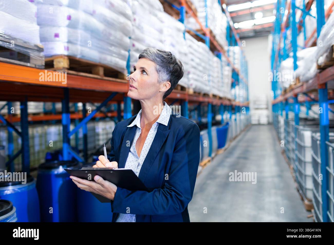 Woman wearing navy blazer with clipboard inspecting pallet racks and ...