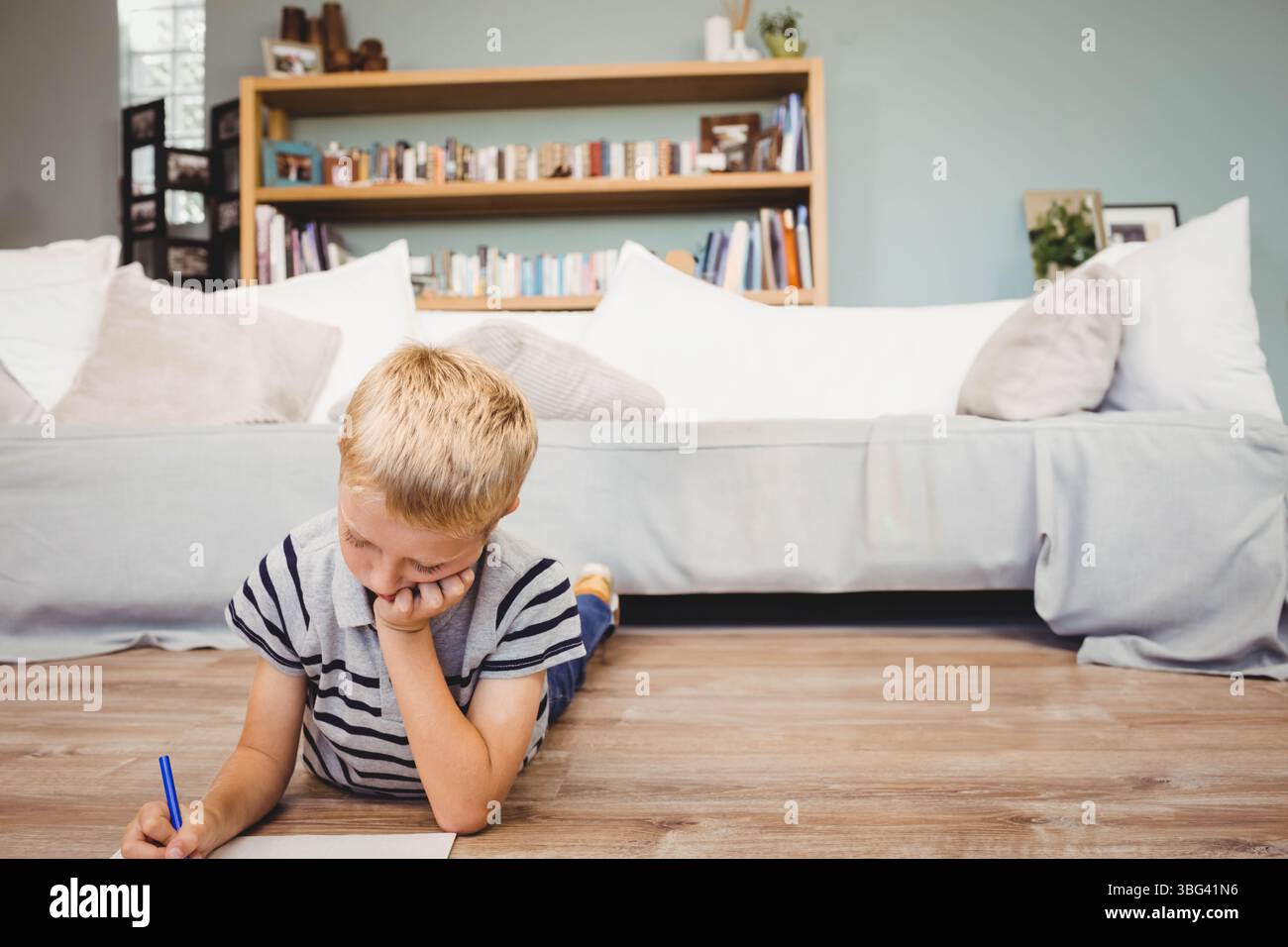 Child writing on wooden floor near grey couch with pillows, blue pen in ...