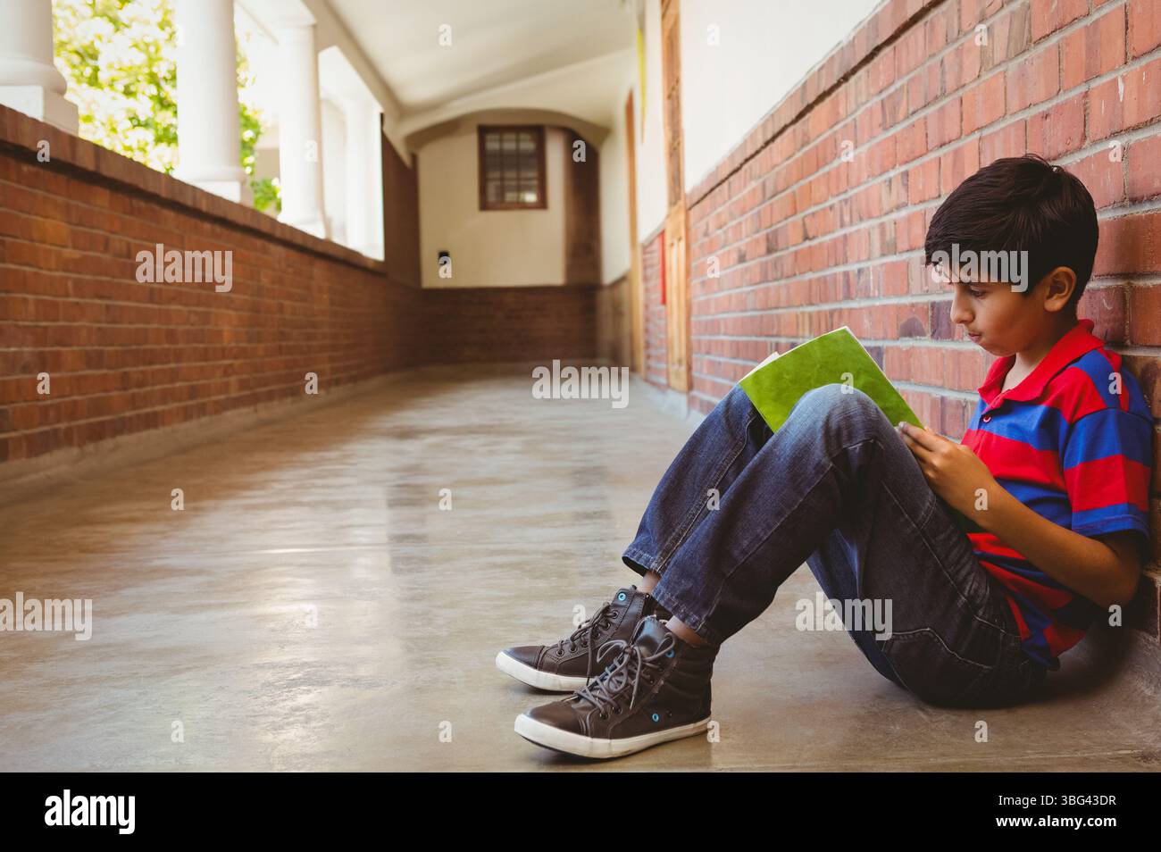 Boy sitting in brick corridor reading green book near white columns ...