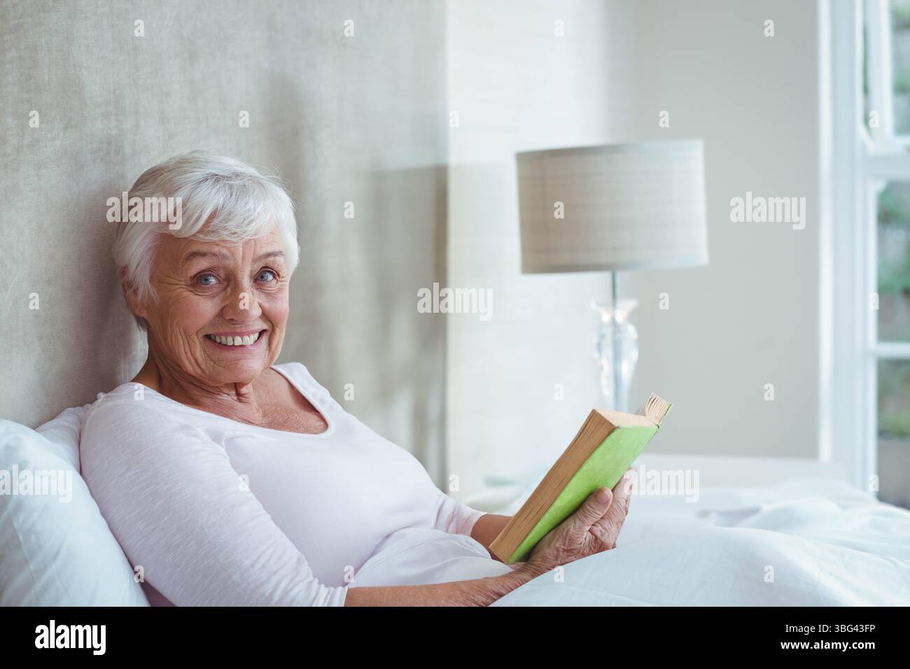 Senior woman sitting up in bed reading open book with green cover by ...