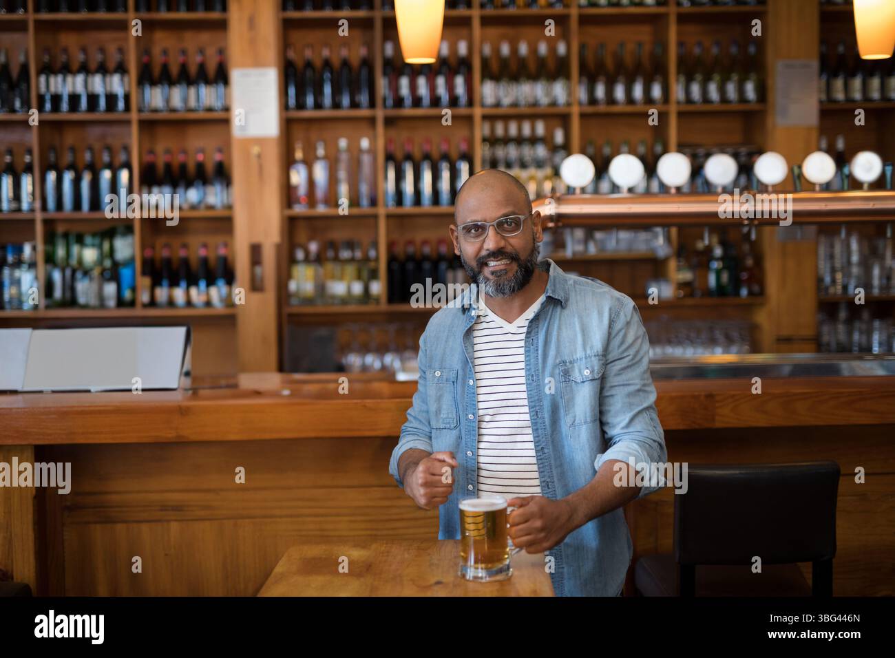Indian man standing at bar counter holding glass beer mug under warm ...