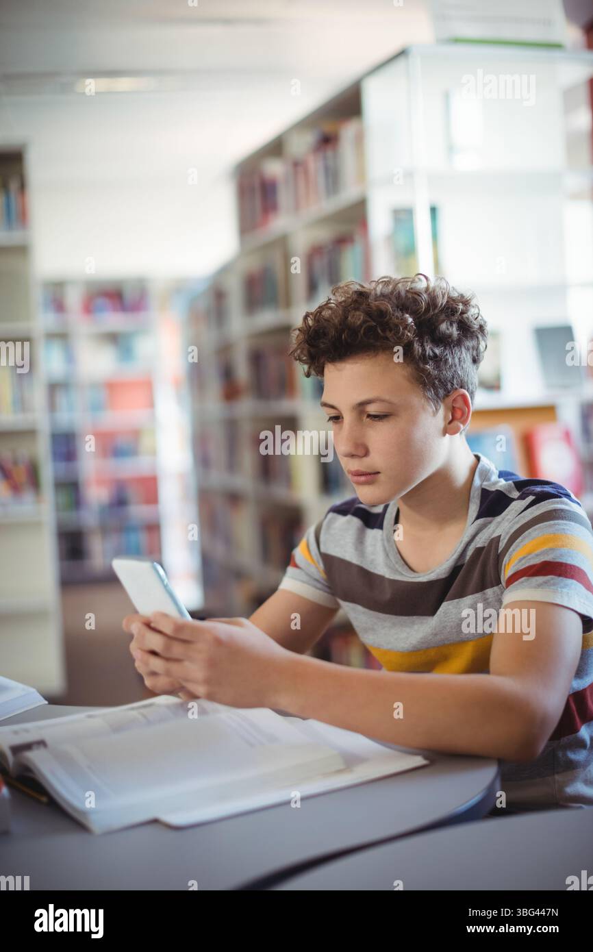 Teenage boy holding smartphone while studying textbook at library round ...