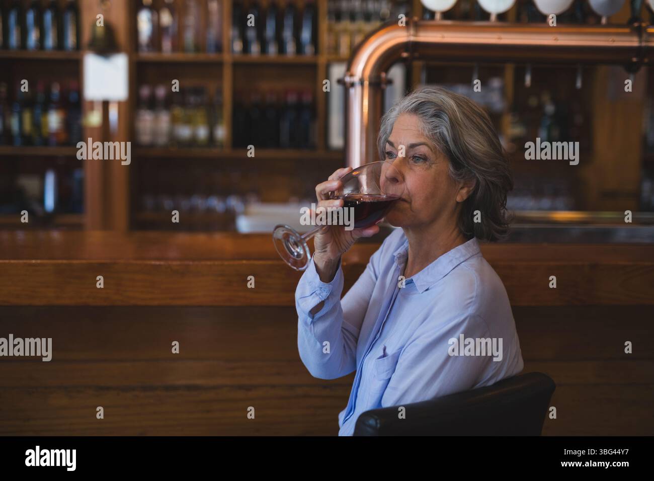 Senior woman raising stemmed wine glass to lips at bar counter with ...