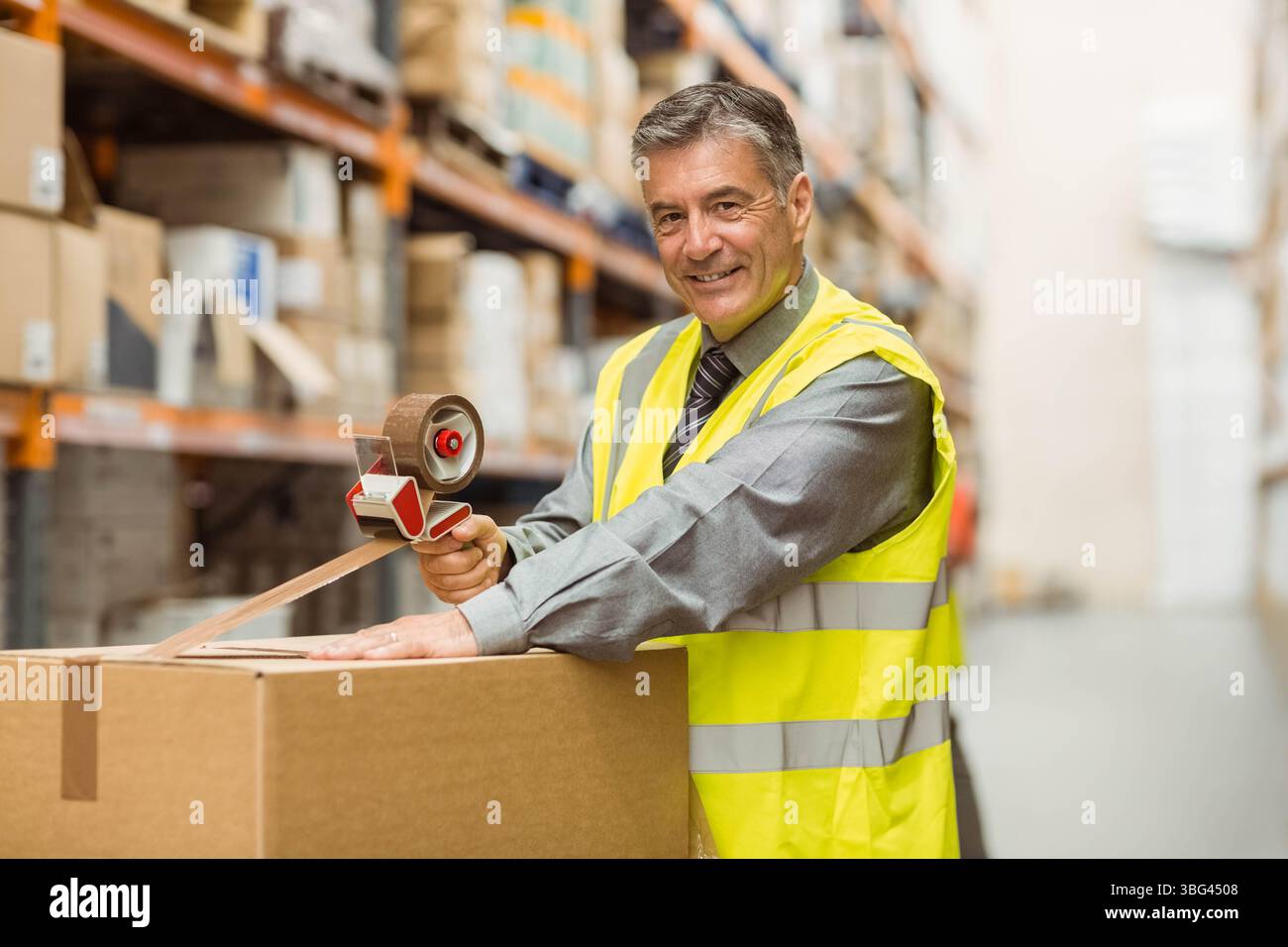 Middle-aged man wearing hi-vis vest sealing cardboard box with tape ...