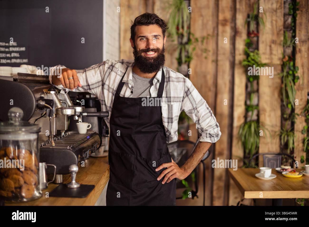 Bearded man wearing black apron leaning on espresso maker at coffee ...