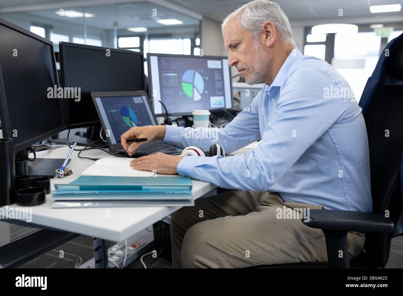 Senior male wearing shirt reviewing pie charts on monitors writing in ...