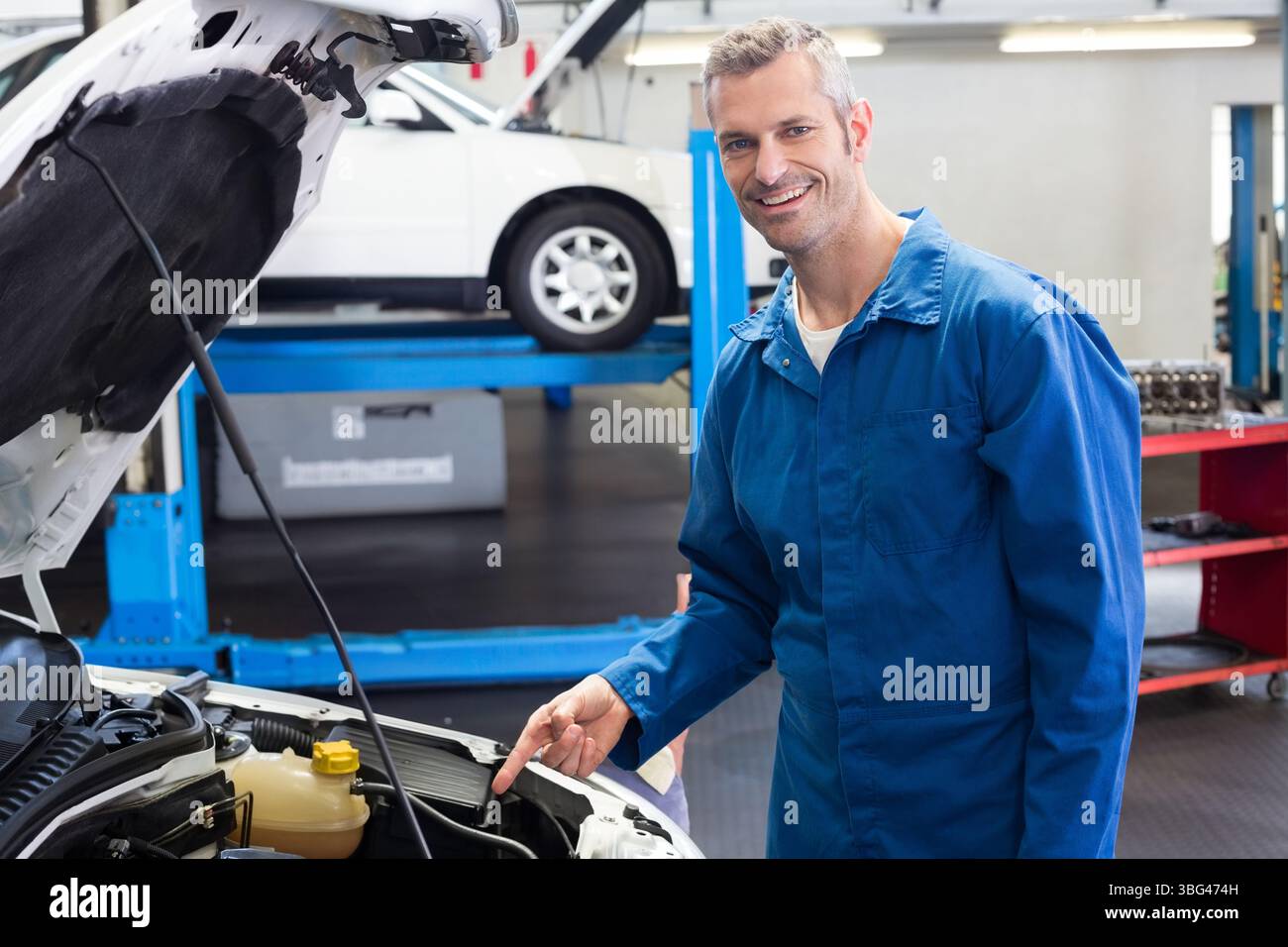 Male mechanic in blue coveralls pointing at coolant reservoir under ...