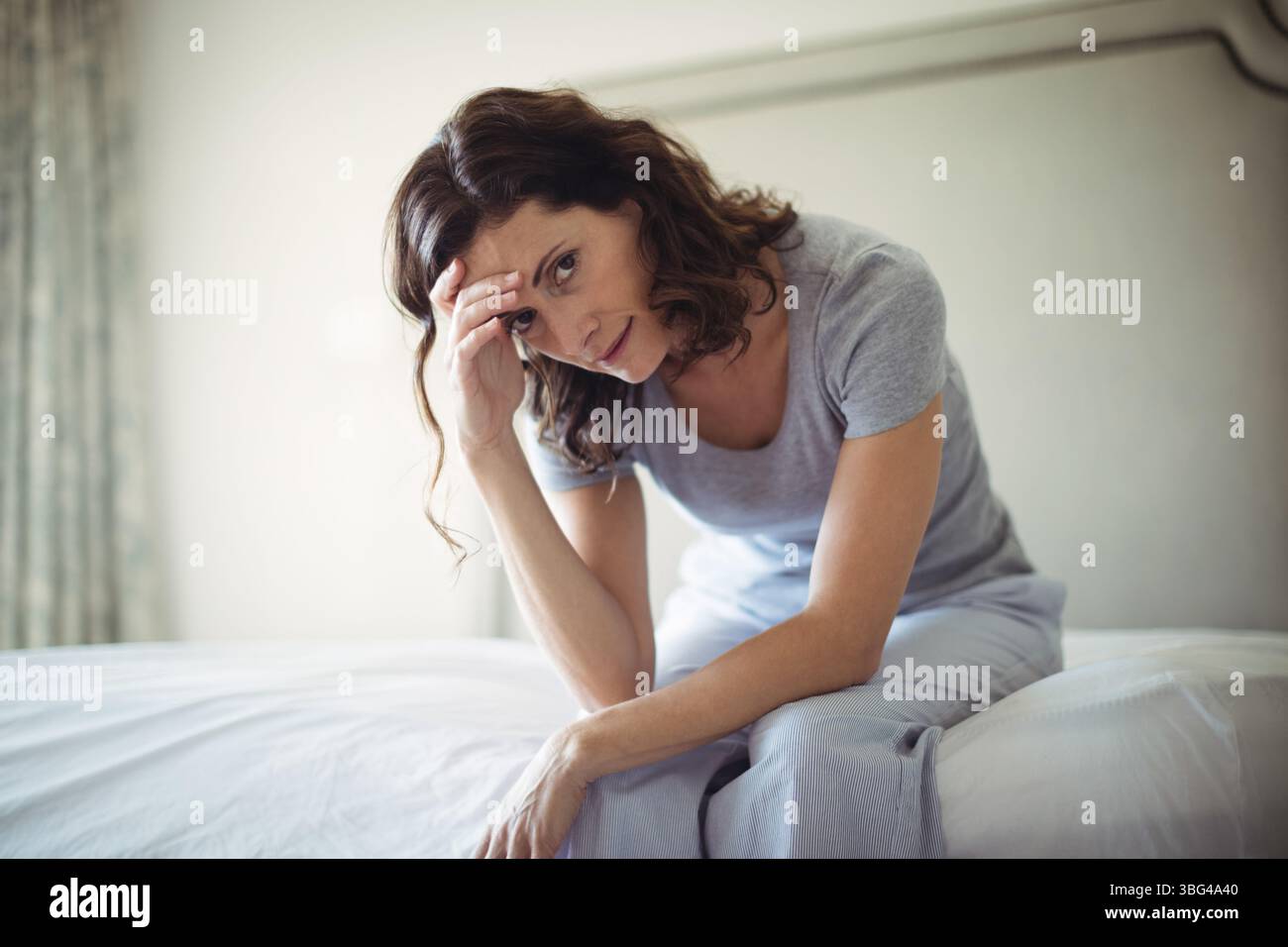 Woman wearing pajama pants sitting on edge of bed in softly lit bedroom ...
