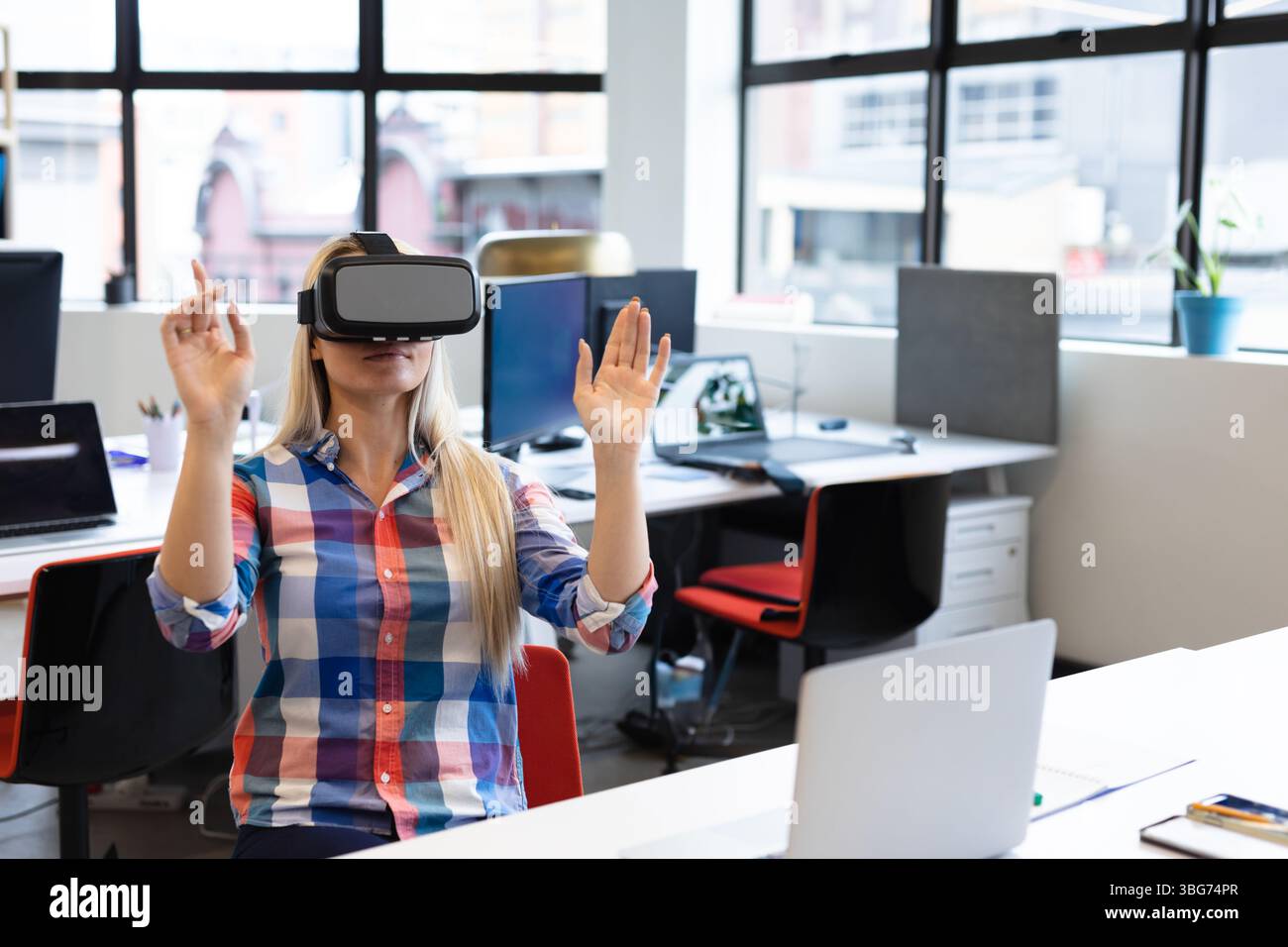 Office worker wearing VR headset raising hands at open plan office desk ...