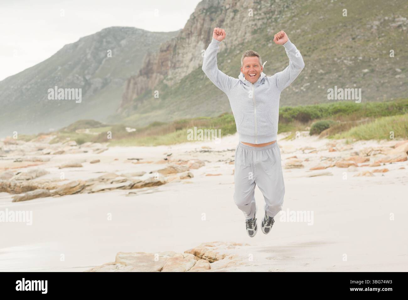 Man jumping mid-air on sandy beach wearing light gray zip-up hoodie ...