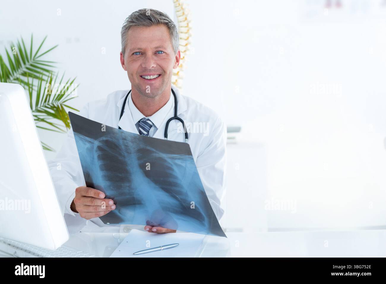 Male doctor examining chest X-ray film at clinic desk with computer ...