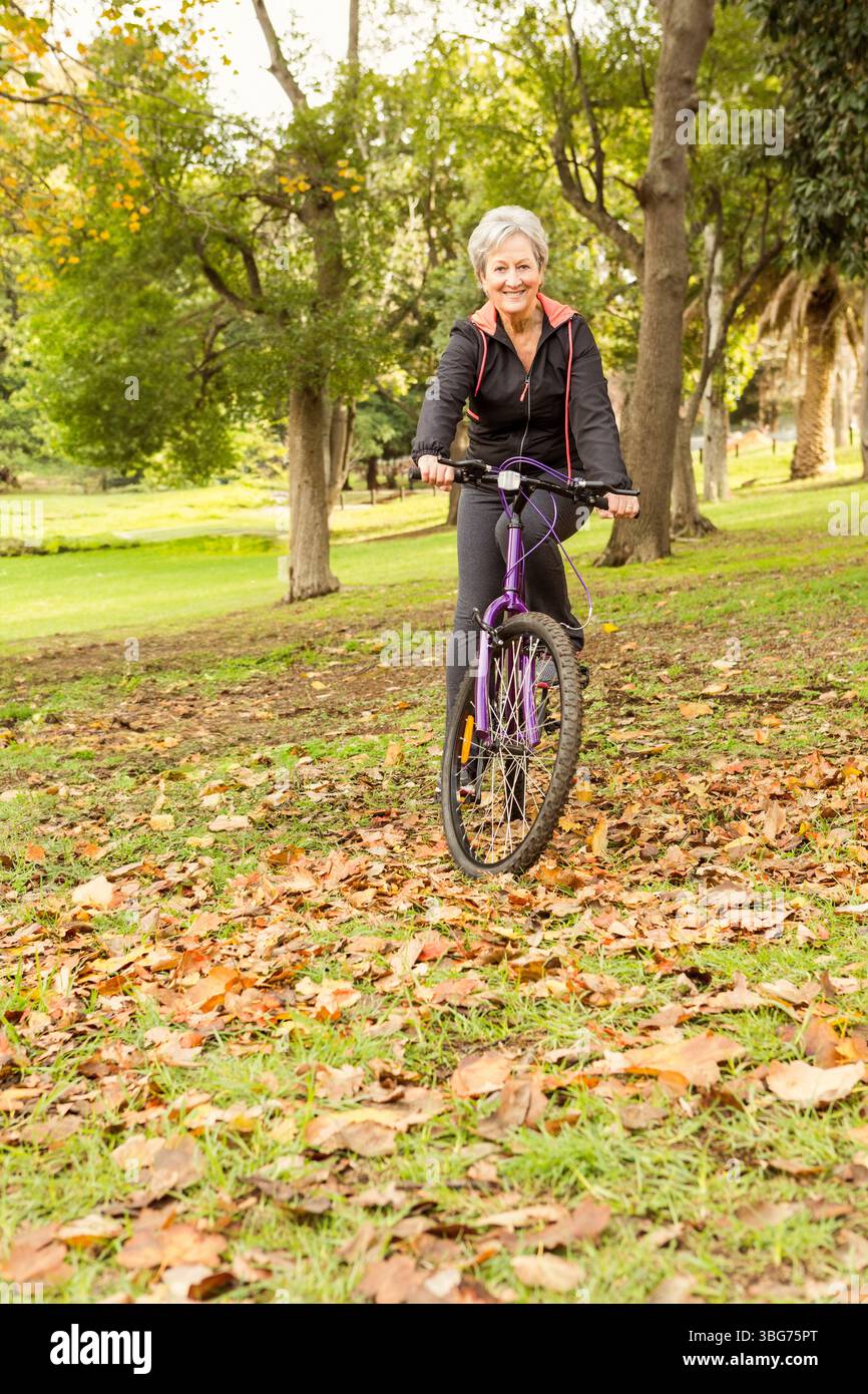 Senior woman cycling through park in autumn on purple bicycle wearing ...
