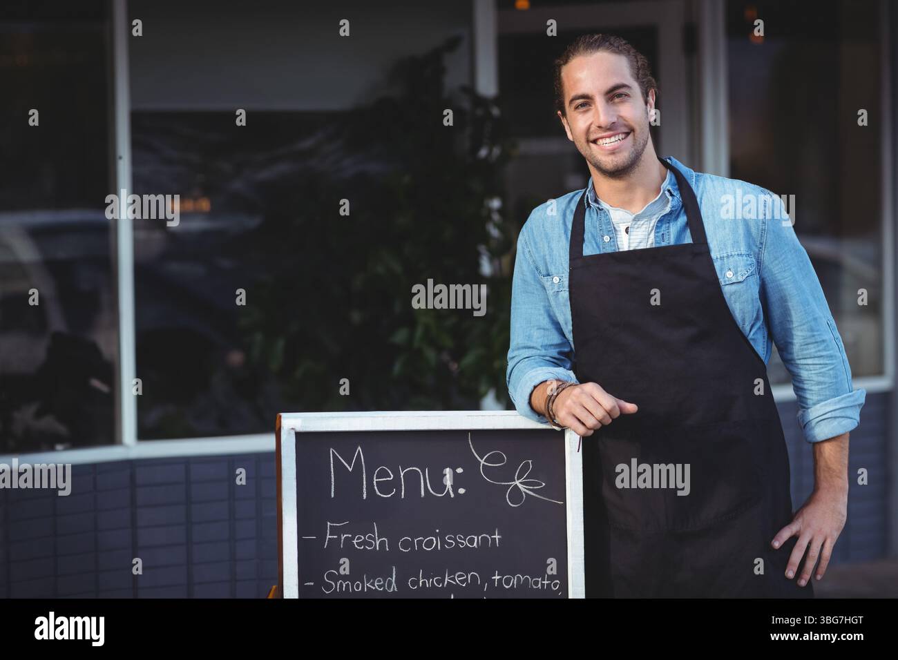 Staff wearing denim shirt and apron leaning on chalkboard outside cafe ...