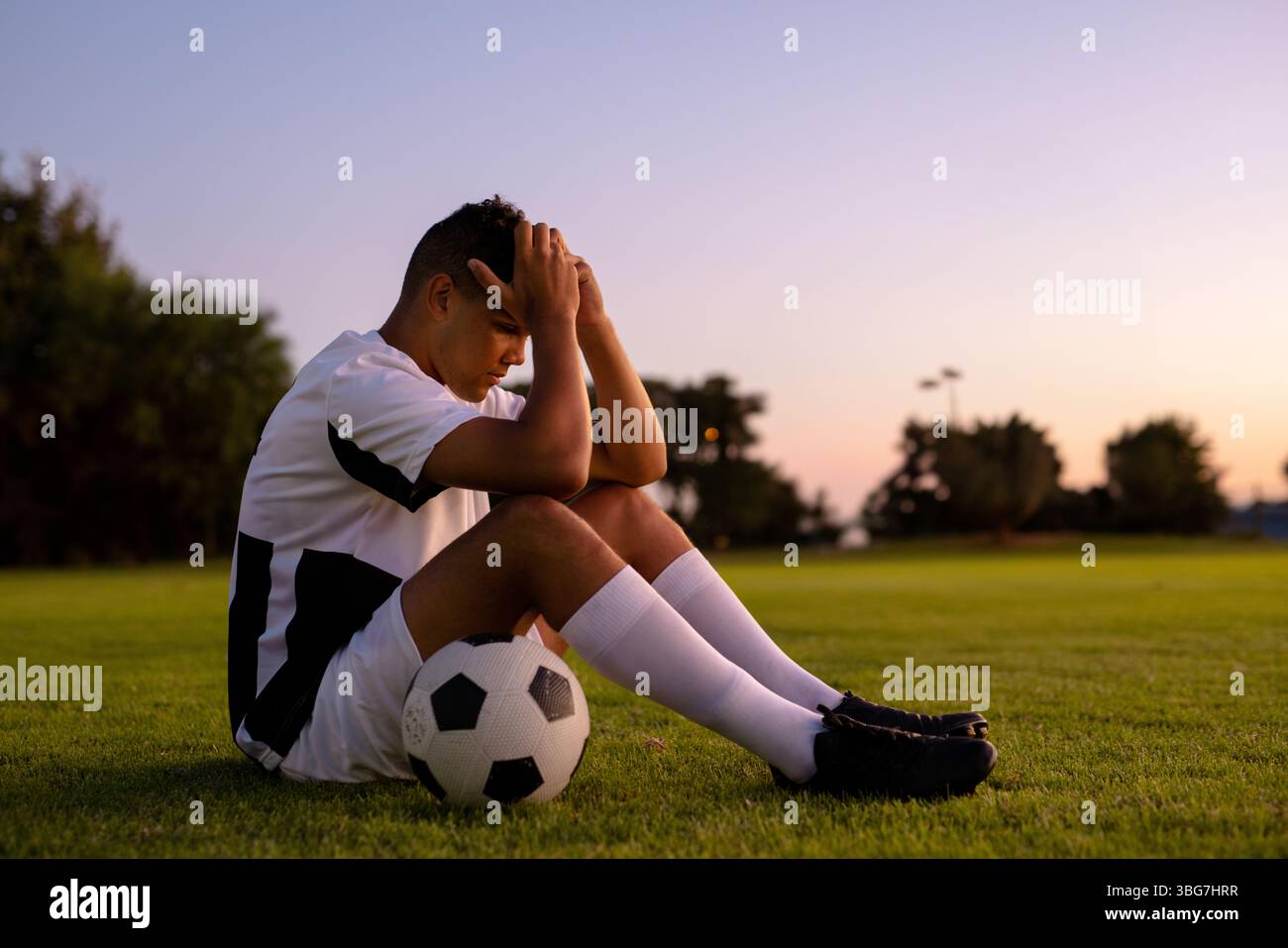 Teen soccer player sitting on grass field at dusk, holding ball under ...