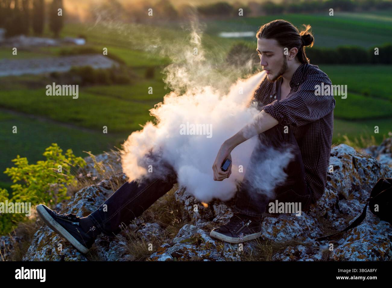 Handsome young man sitting on a rocks and smoking electronic cigarette ...