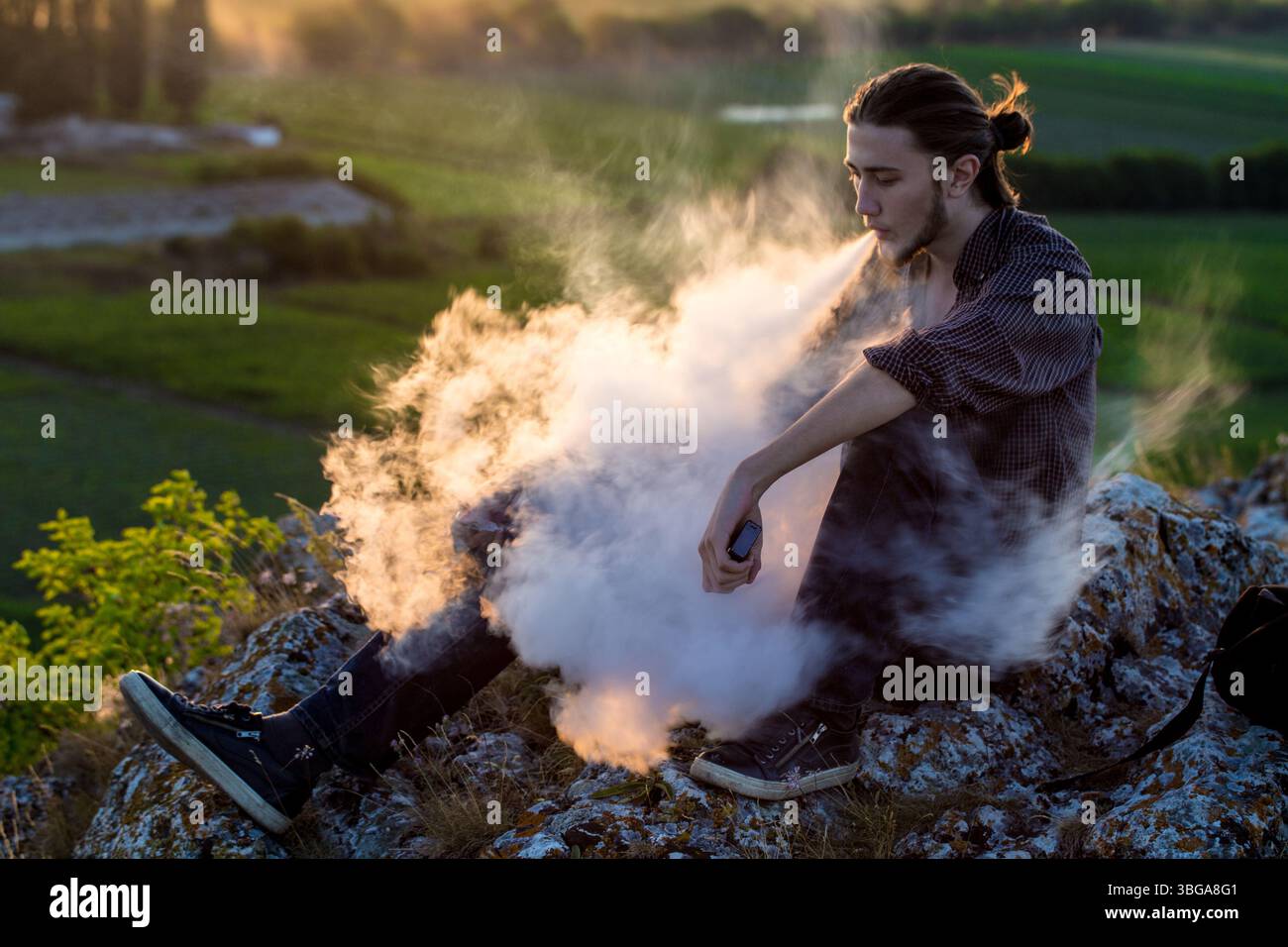Handsome young man sitting on a rocks and smoking electronic cigarette ...