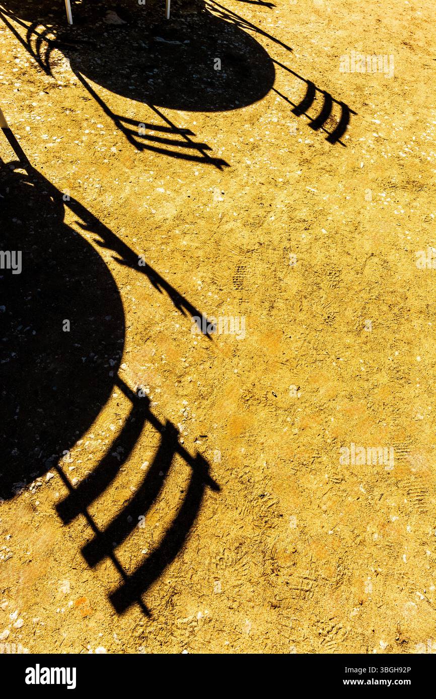 Shadows cast by chairs and tables stretch across a sunlit ground ...
