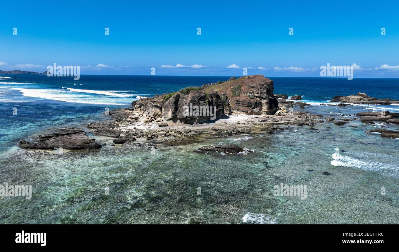 Rocky outcrop rises from shallow coral reefs, where waves break and the ...